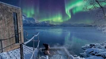 Pessoa em banheira de hidromassagem ao ar livre, com vista para a aurora boreal.