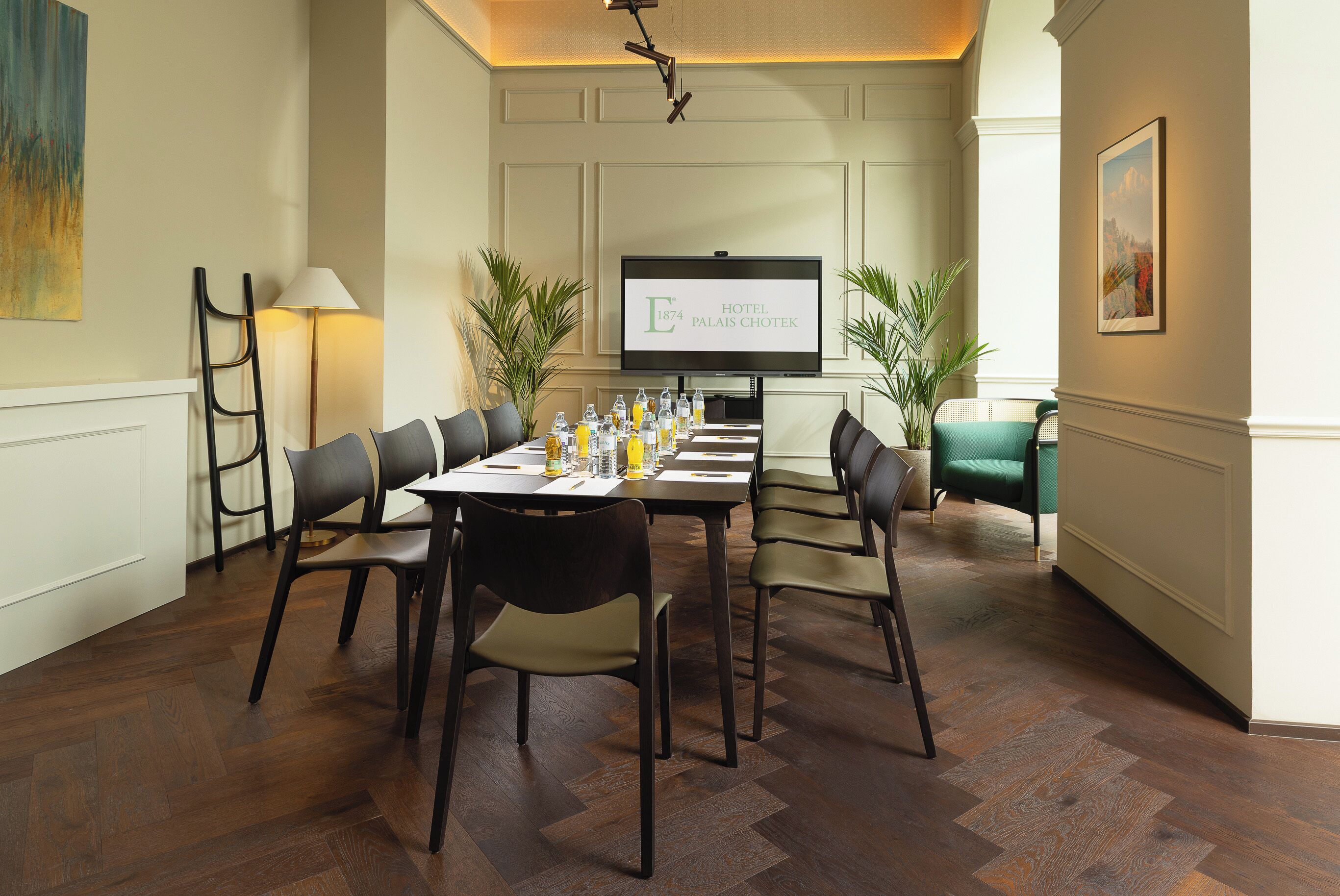 Uma sala de conferências de hotel com uma mesa comprida, cadeiras, uma tela de TV e um quadro na parede.