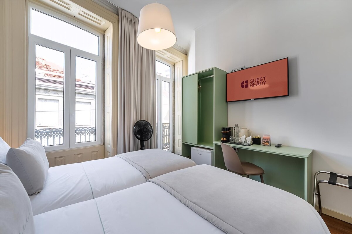Quarto de hotel com duas camas, uma escrivaninha, uma cadeira, um ventilador, uma televisão e uma janela com cortinas.