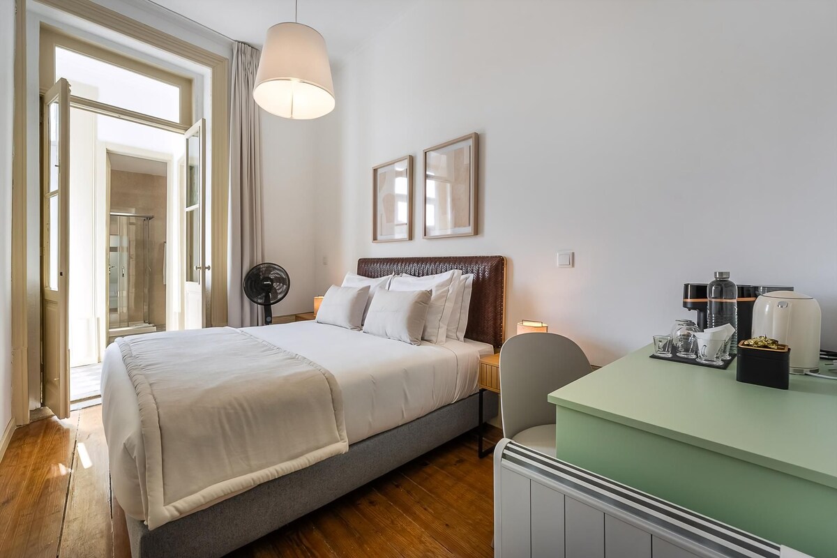 Quarto com cama, mesa de cabeceira e escrivaninha com chaleira e chávenas.