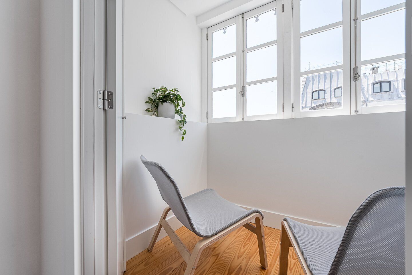 Quarto com duas cadeiras, uma planta em vaso e janela com vista para edifícios.