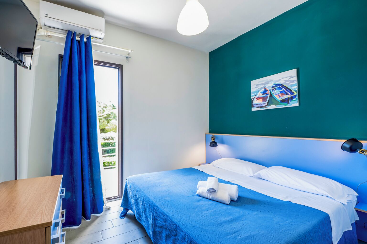 Quarto de hotel com cama, escrivaninha, janela com cortinas, um quadro na parede e uma luminária no teto.
