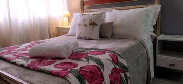 Cama bem arrumada com roupa de cama floral, travesseiro branco com desenho de pássaro e toalha branca dobrada.