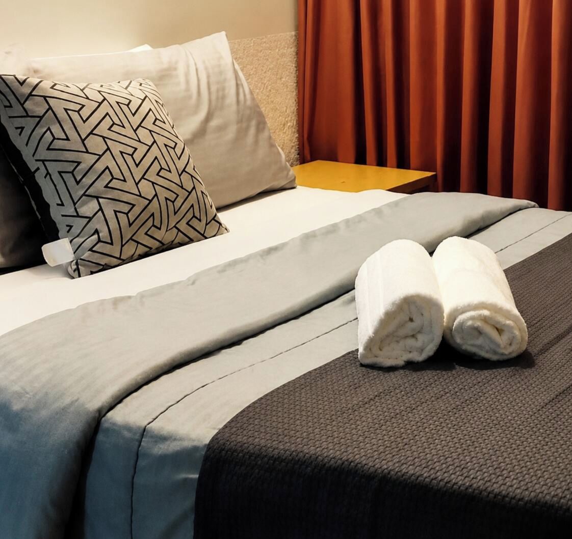 Cama bem arrumada com travesseiro estampado, toalha dobrada e uma pequena mesa de cabeceira de madeira.