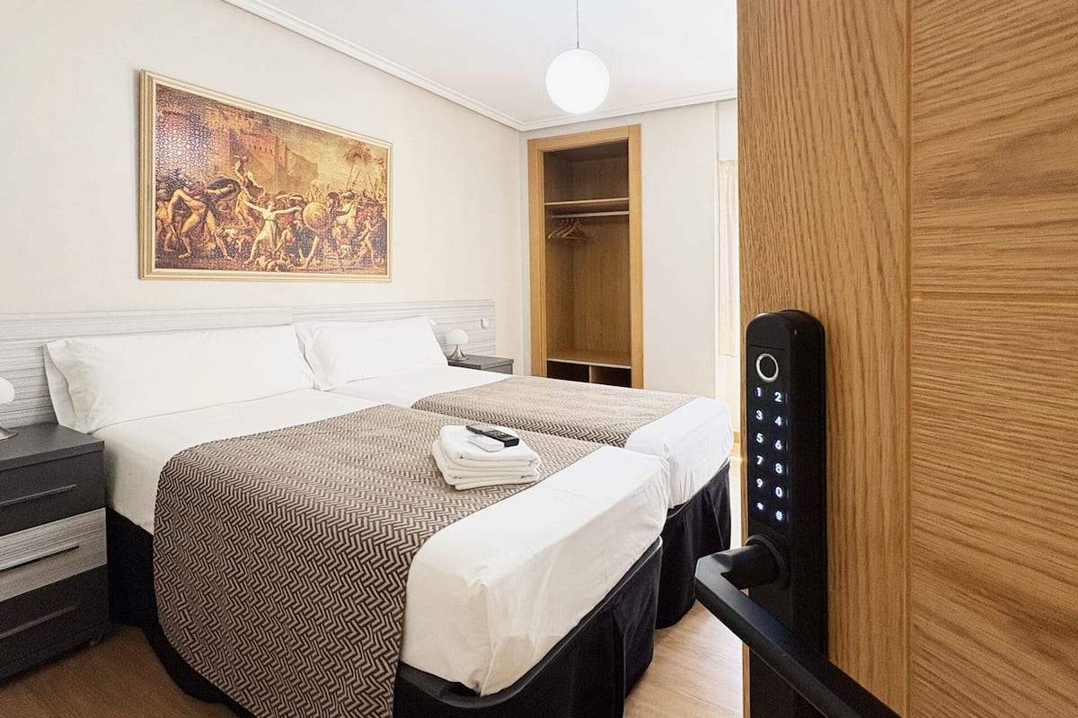 Quarto de hotel com cama, mesa de cabeceira, quadro, guarda-roupa e porta com fechadura eletrônica.