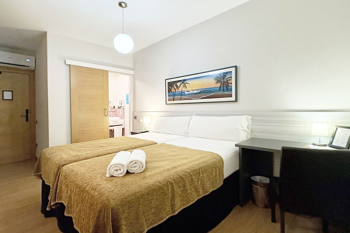 Quarto de hotel com cama, escrivaninha, cadeira, quadro de uma praia e porta para o banheiro.