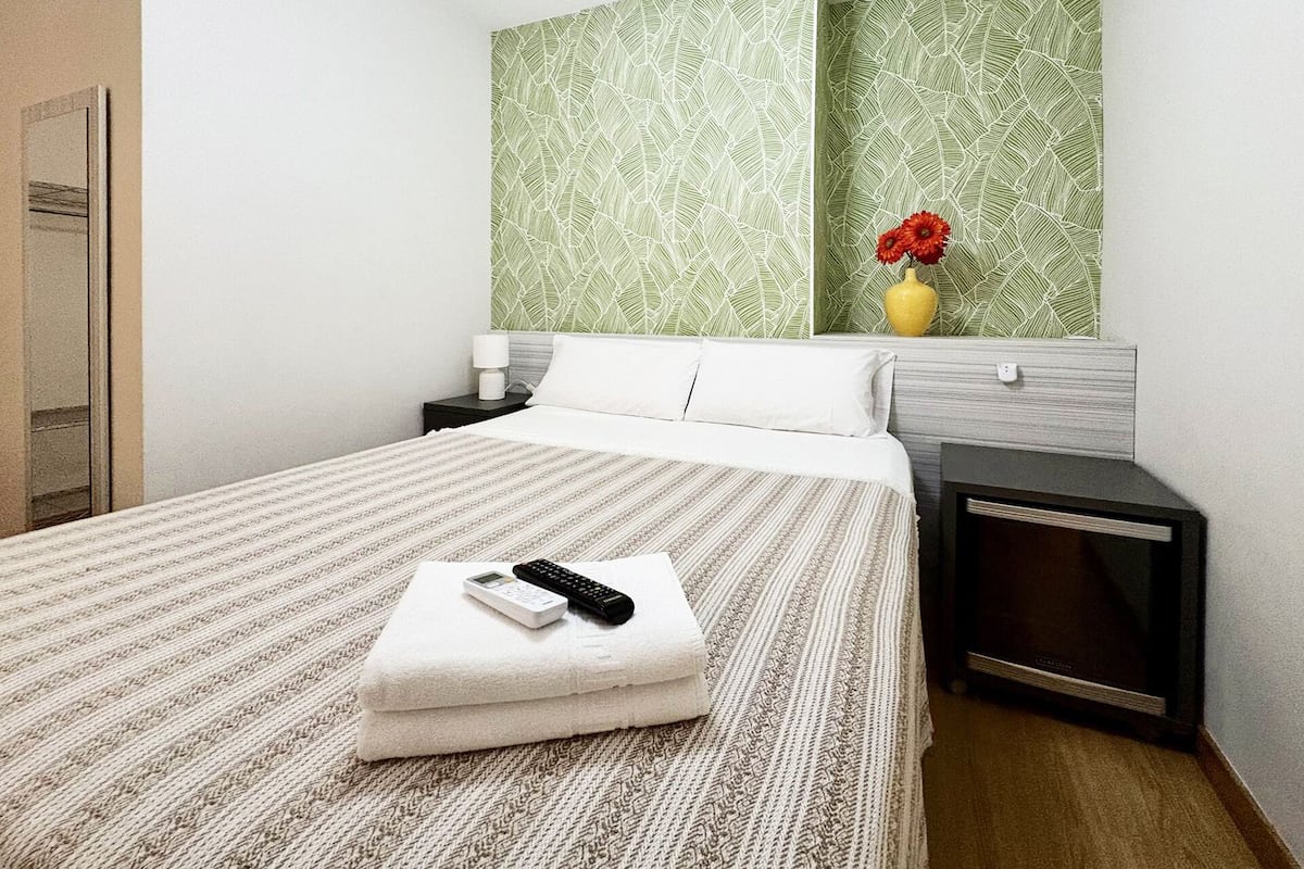 Quarto de hotel com cama, mesa de cabeceira, luminária, controle remoto e um vaso com flores.