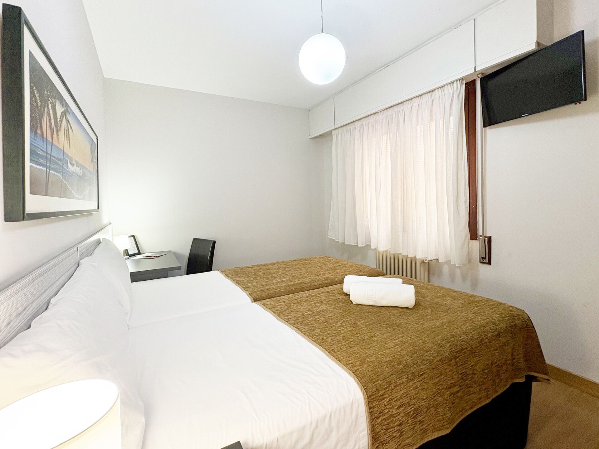 Quarto de hotel com cama, escrivaninha, cadeira, televisão e janela com cortinas.