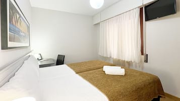 Quarto de hotel com cama, escrivaninha, cadeira, televisão e janela com cortinas.