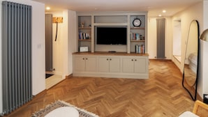 Uma sala de estar moderna com uma televisão de tela plana grande, piso de madeira e estantes embutidas com livros e objetos decorativos.