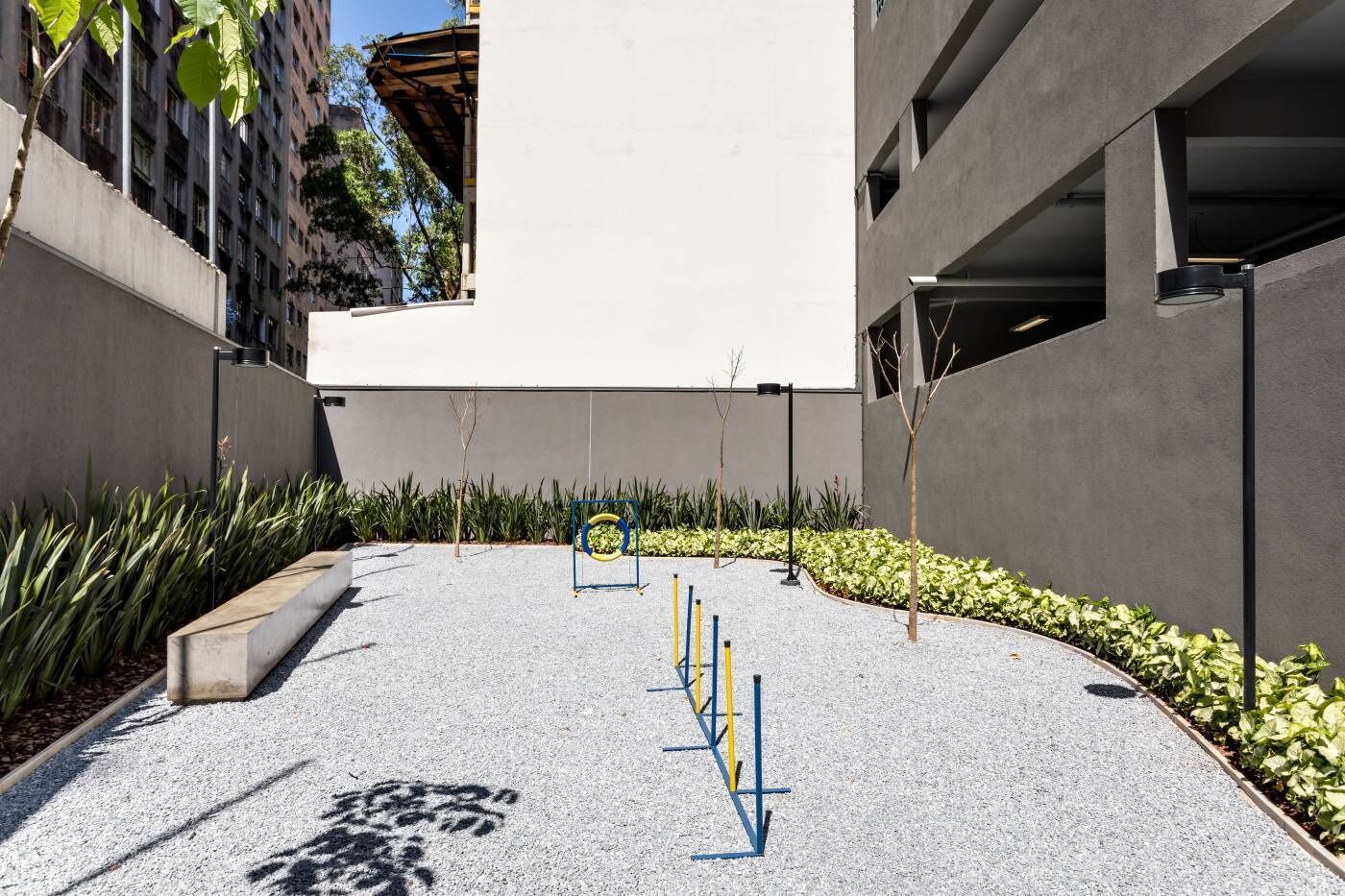 Um pátio com piso de concreto, uma pequena área de jardim com plantas e uma estrutura metálica com postes amarelos e azuis.