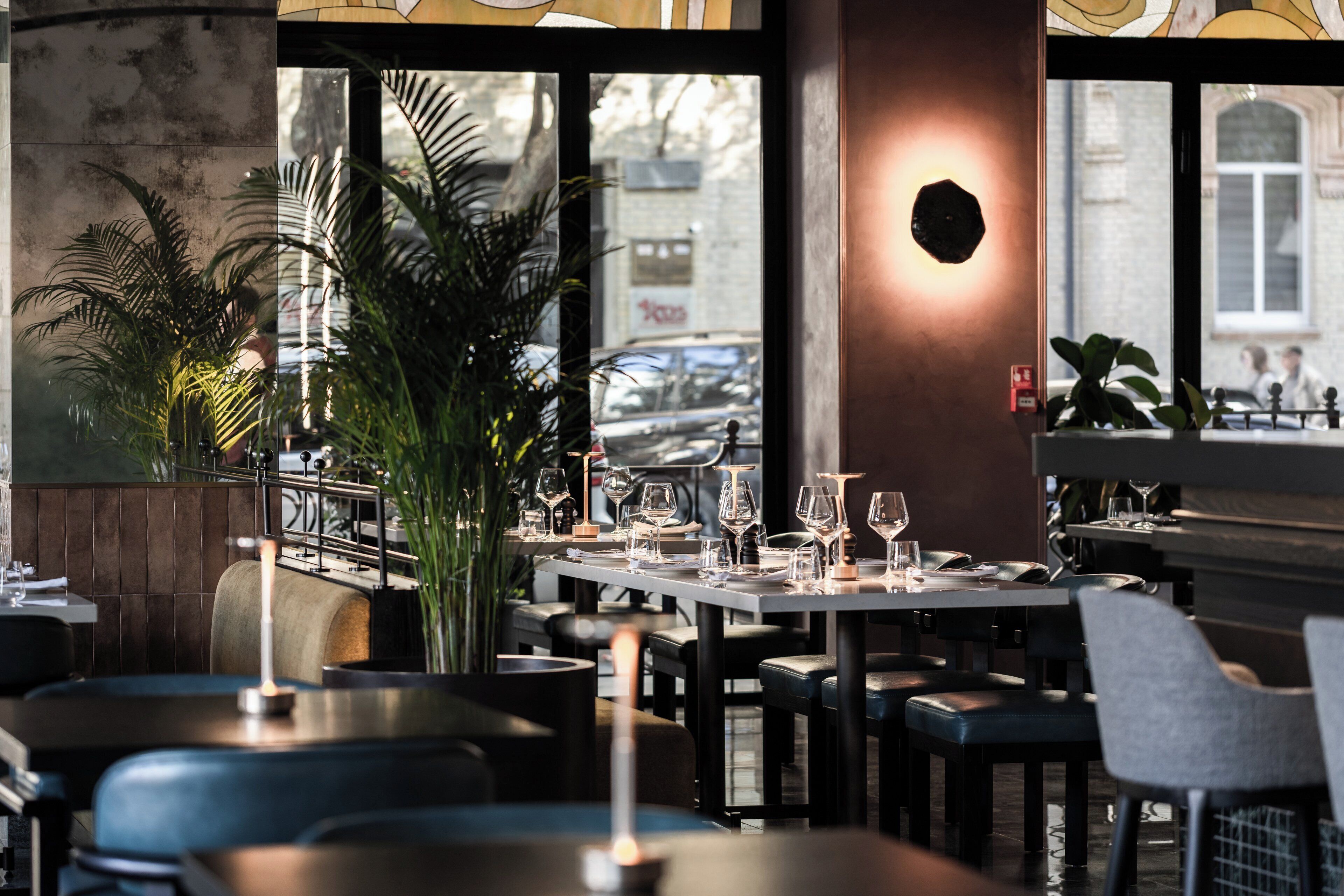 Um restaurante com mesas preparadas para refeições, grandes janelas e plantas em vasos.