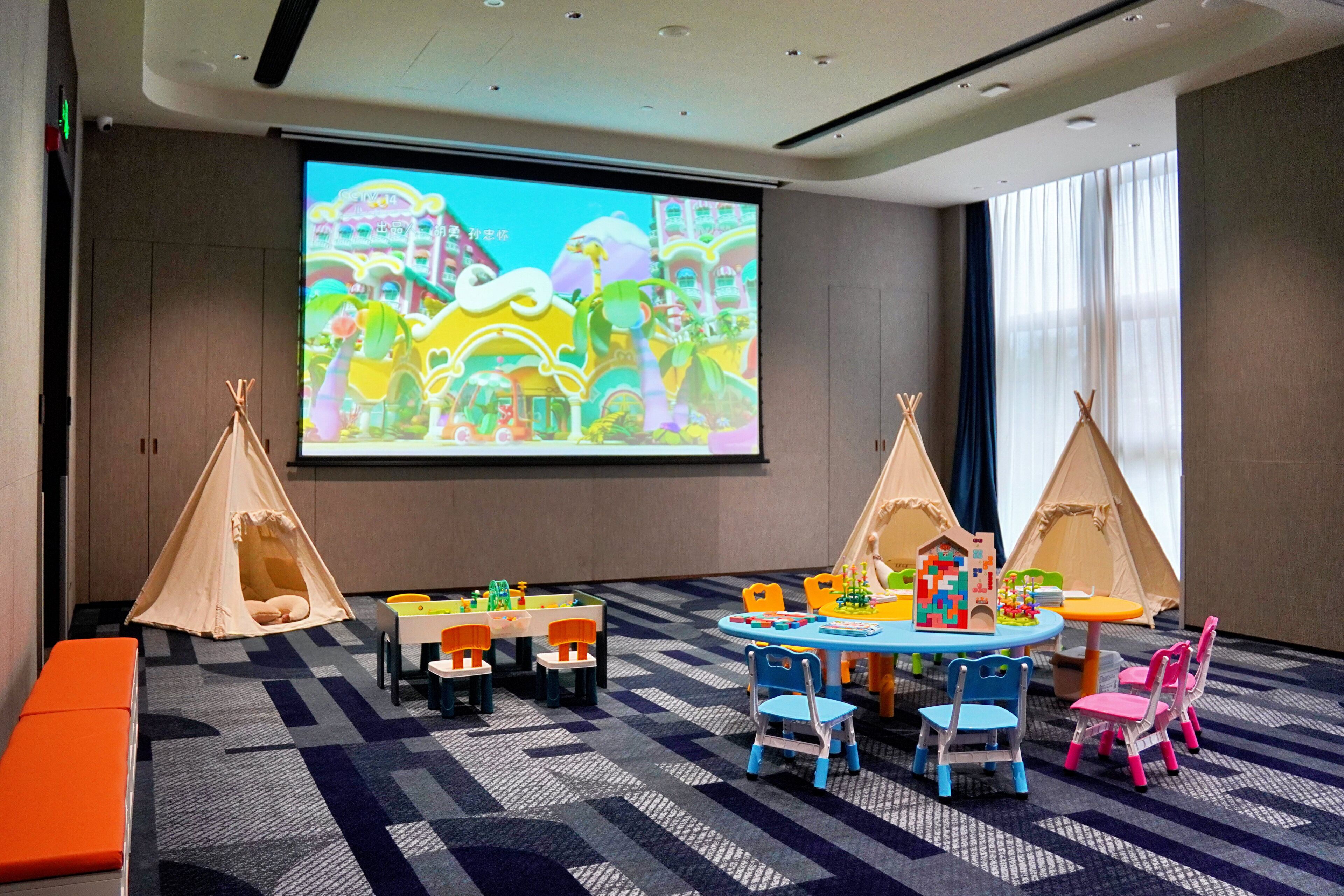 Uma sala de conferências com uma tela grande exibindo uma animação colorida, uma mesa redonda com cadeiras e dois pequenos tipi.