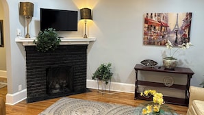 Uma sala de estar com lareira, televisão de tela plana, luminárias decorativas, um quadro e uma mesa de centro de vidro com flores.