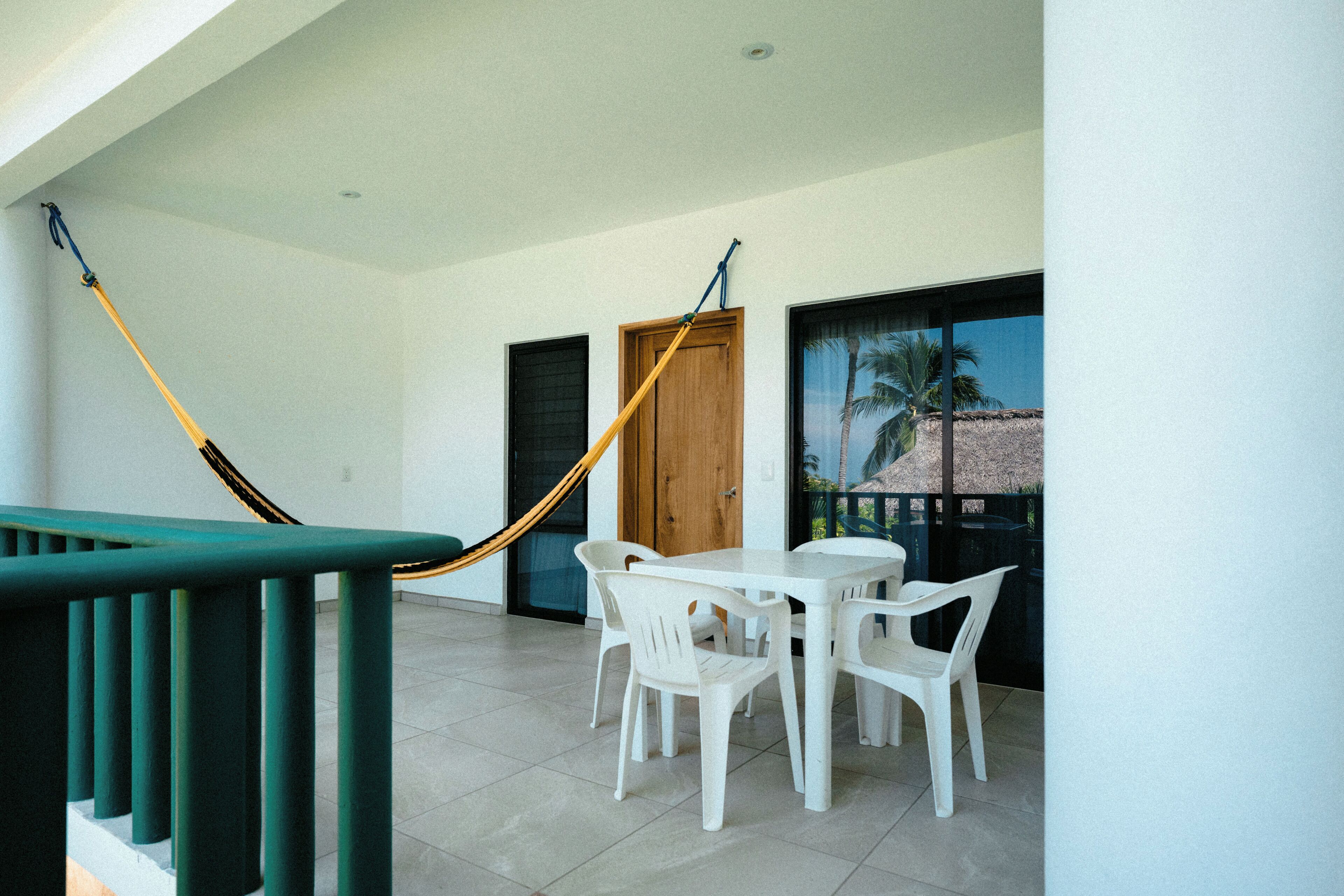Uma varanda com mesa e cadeiras, uma porta de madeira e vista para palmeiras.