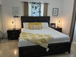 Quarto com cama, duas mesas de cabeceira, painel de cabeceira, janela com cortinas e decorações nas paredes.