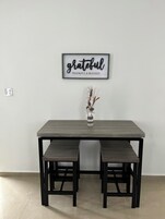 Uma mesa de jantar com duas bancos, uma placa na parede acima e um vaso com plantas secas.