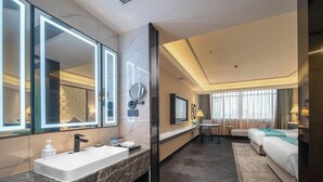 Quarto de hotel moderno com toucador em mármore, espelho redondo, cama grande, sofá e mesa pequena.