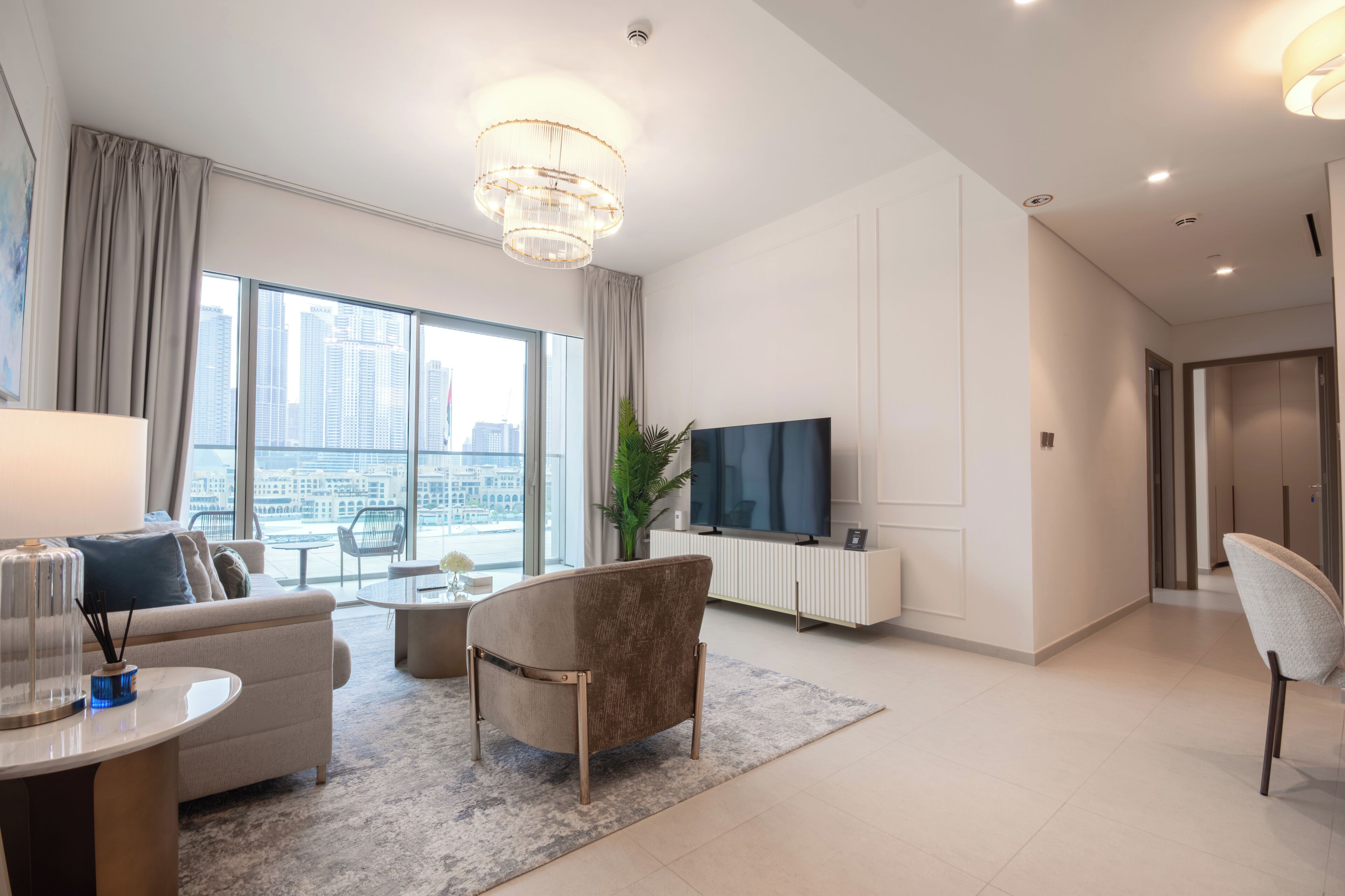 Uma sala de estar moderna com sofá, poltrona e mesa de centro. Uma televisão está instalada na parede, e há uma janela grande com vista para edifícios.