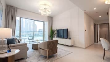 Uma sala de estar moderna com sofá, poltrona e mesa de centro. Uma televisão está instalada na parede, e há uma janela grande com vista para edifícios.