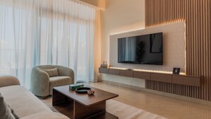 Uma sala de estar moderna com uma TV de tela plana na parede, um sofá confortável, uma mesa de centro e uma janela grande com cortinas leves.