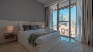Quarto de hotel moderno com uma cama grande, mesa de cabeceira com luminária e vista para a cidade através de amplas janelas.