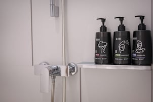 Casa de banho com chuveiro, sanita e uma prateleira com três frascos pretos de champô, condicionador e gel de banho.