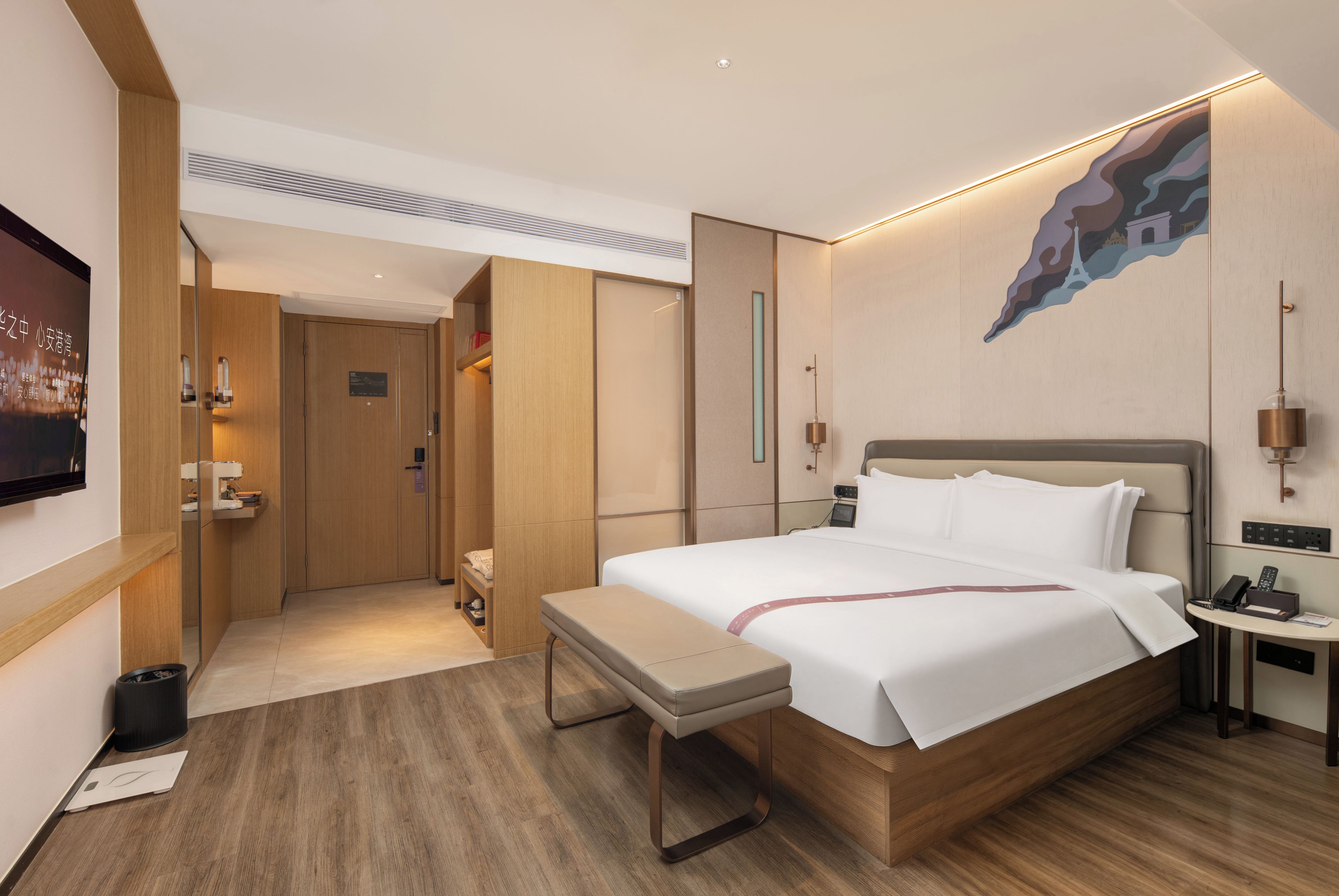 Quarto de hotel com uma cama grande, um banquinho, uma escrivaninha, televisão na parede e um quadro.