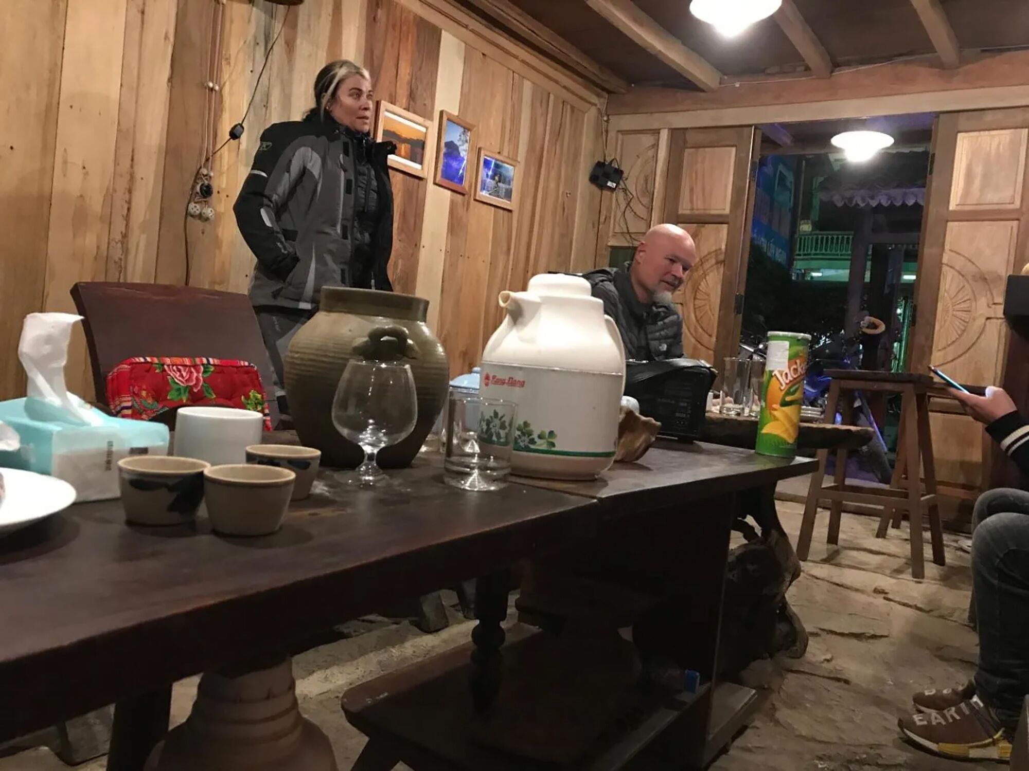 Interior rústico de madeira com uma mesa posta para refeição, apresentando uma jarra de cerâmica grande, uma cafeteira e várias xícaras.
