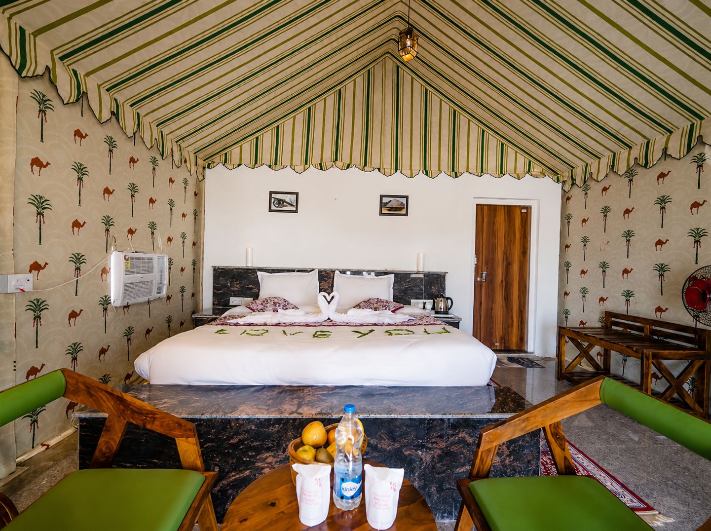 Quarto de hotel com cama, mesa com frutas e uma garrafa, e uma cadeira de madeira.