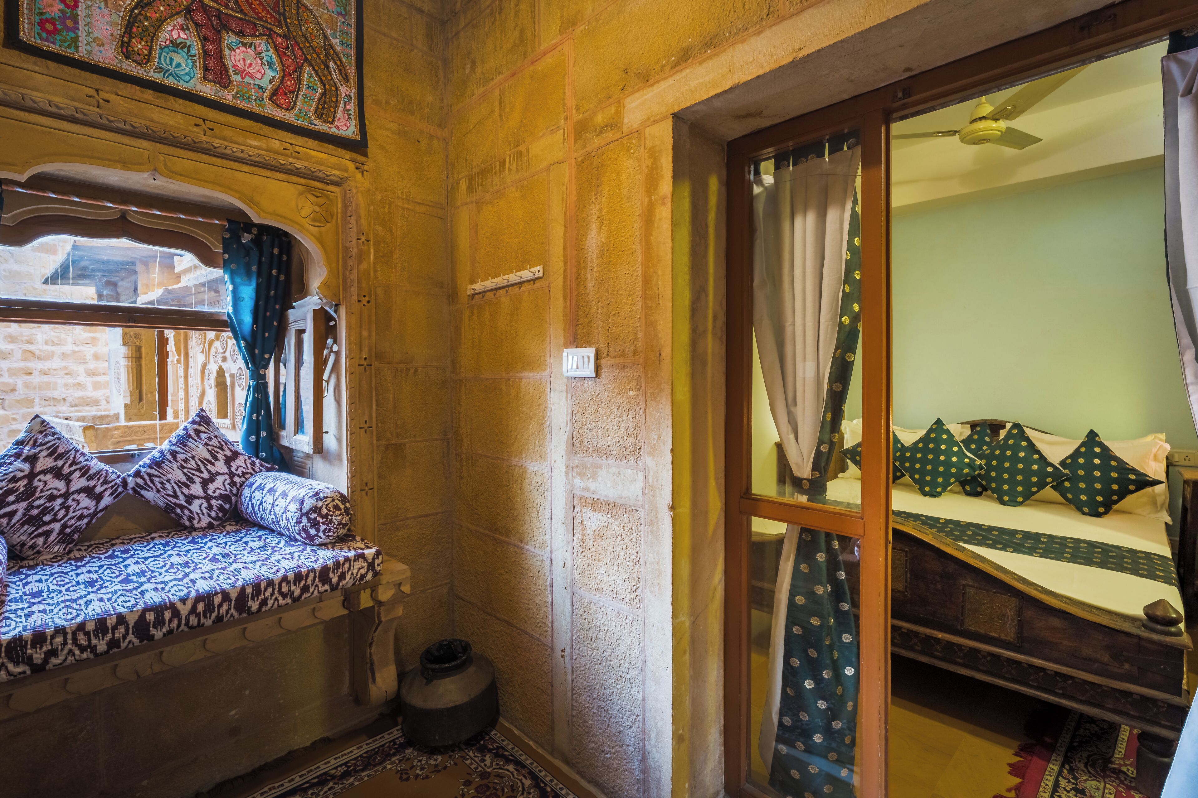 Quarto tradicional com uma divisória baixa, janela com cortinas estampadas, cama com almofadas verdes e um pequeno tapete.