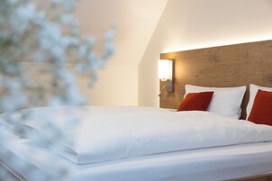 Cama bem arrumada com lençóis brancos e duas almofadas vermelhas em quarto com cabeceira de madeira e luminária fixada na parede.