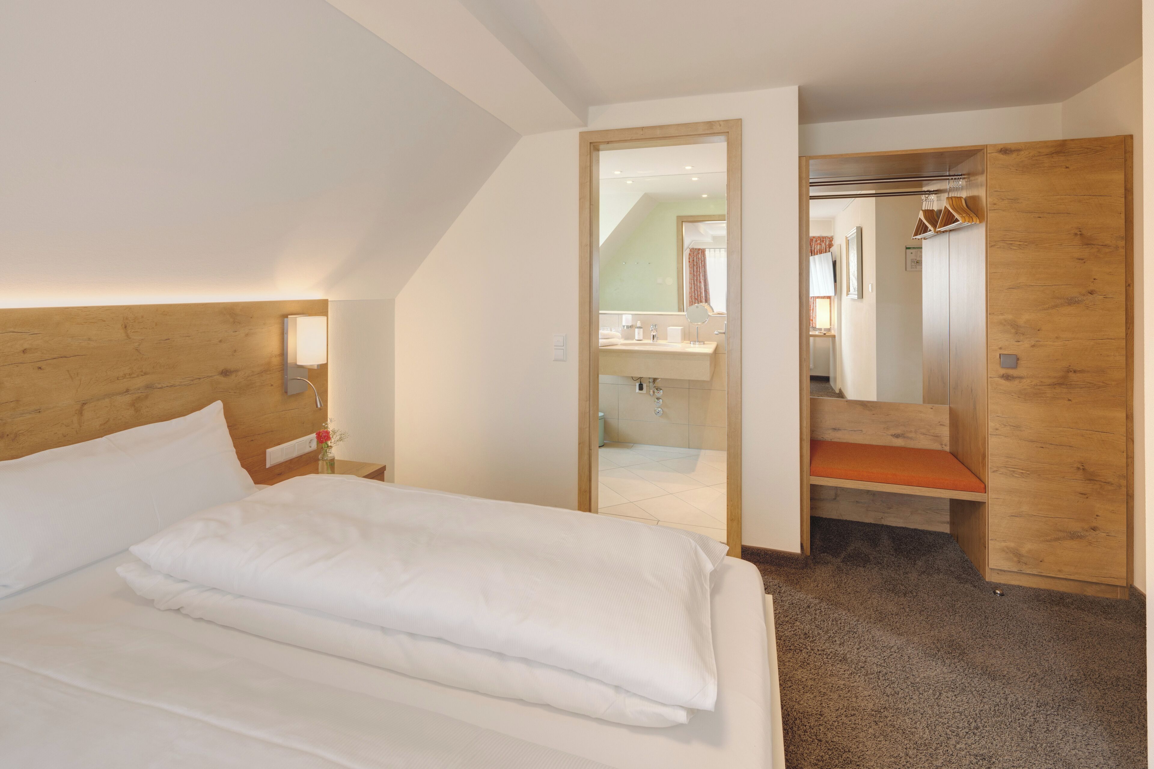 Quarto de hotel com cama, cabeceira de madeira, mesa de cabeceira com abajur, armário e banheiro visível por uma porta aberta.
