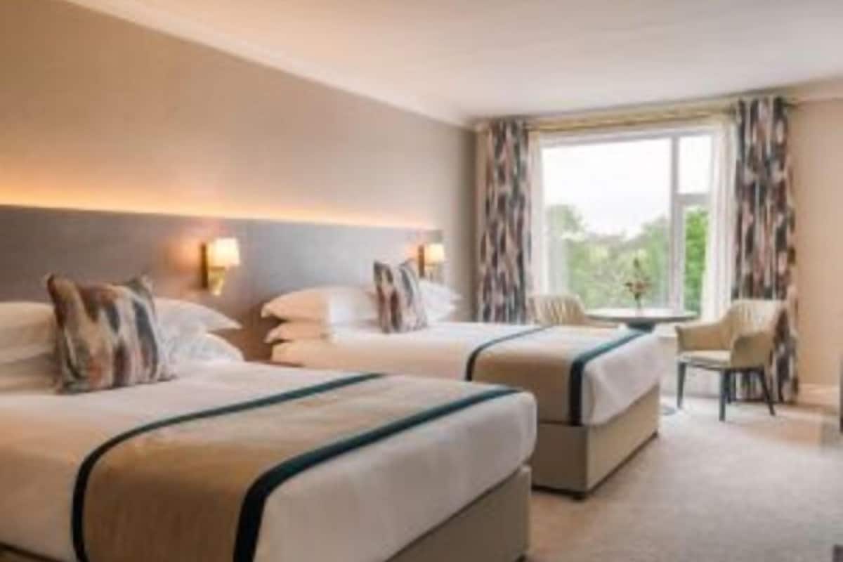 Quarto de hotel com duas camas, área de estar e janela com cortinas.