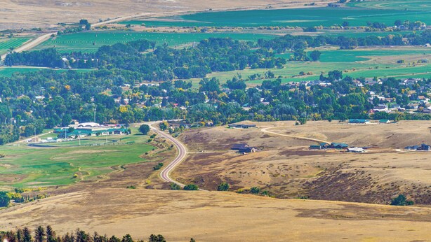 Paisagem rural com estrada sinuosa, campos verdes e um pequeno conjunto de casas.