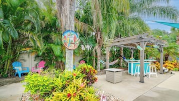 Uma área externa tropical com um pergolado de madeira, uma rede, uma escultura de pássaro colorida e um espaço de bar com banquetas.