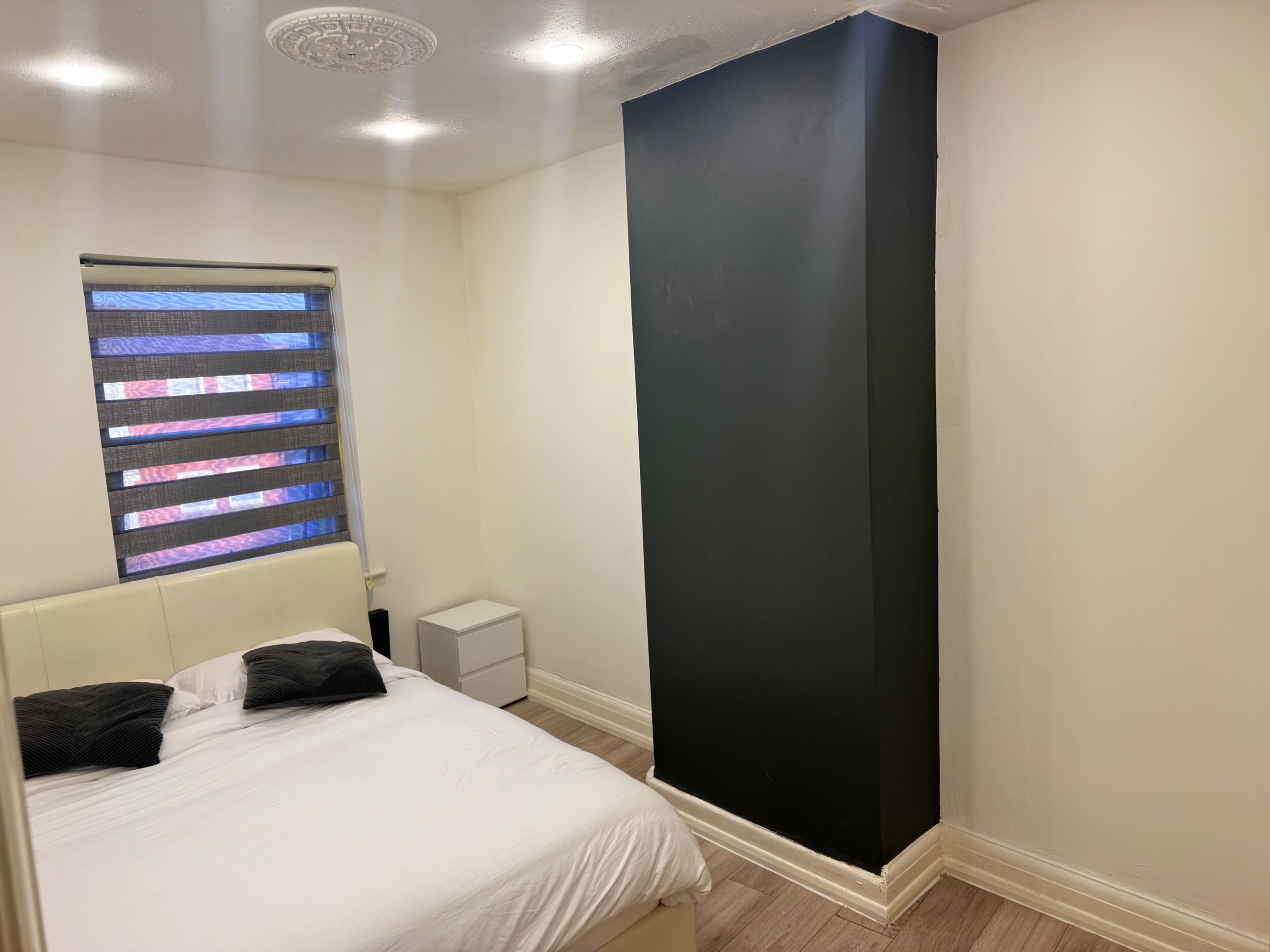 Quarto com cama, mesa de cabeceira, janela com persianas e uma parede de destaque escura.