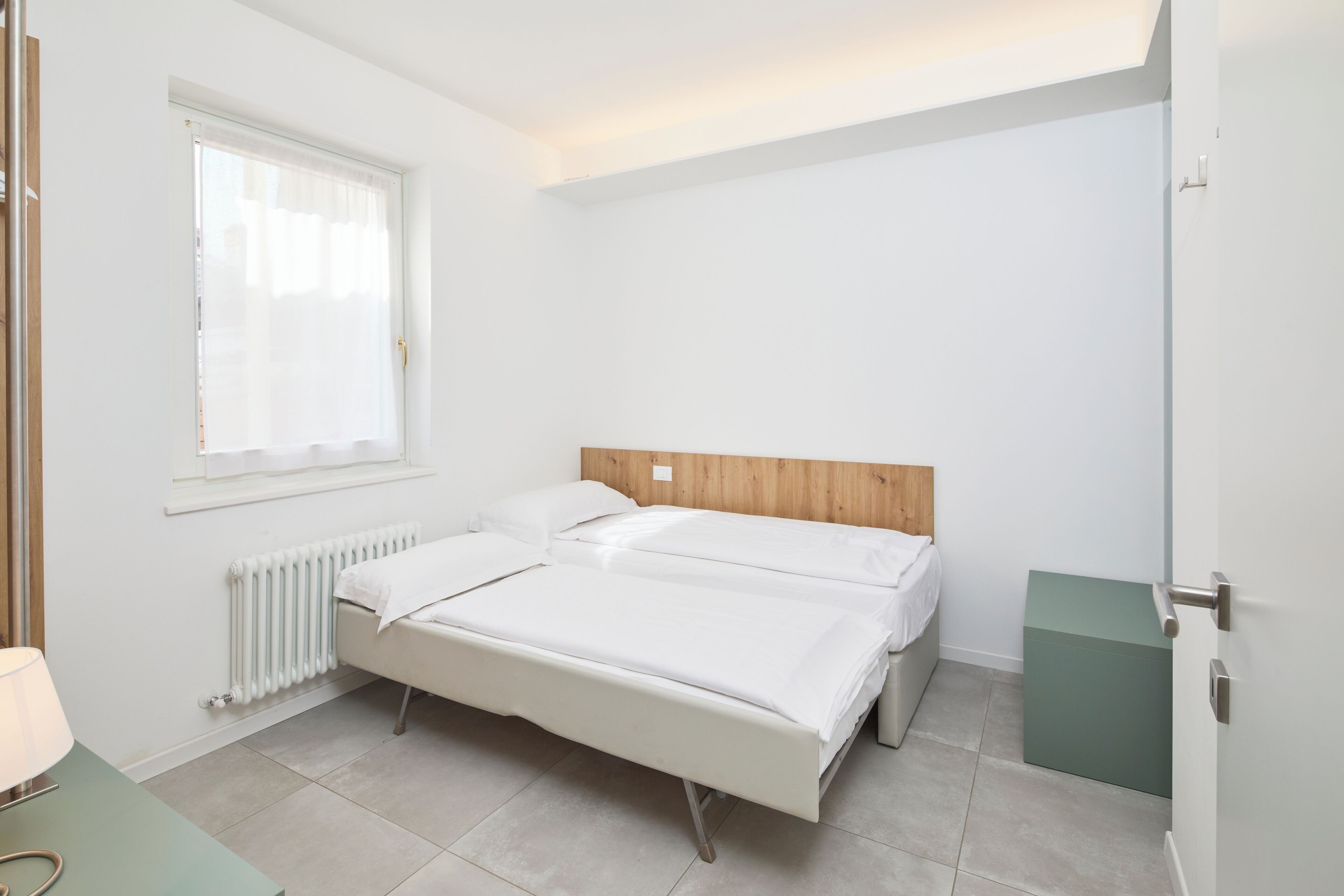 Quarto moderno com cabeceira de madeira, duas camas, um radiador e uma janela com cortinas leves.