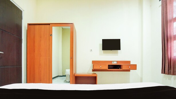 Quarto de hotel com uma cama, um guarda-roupa de madeira, uma pequena mesa de madeira, uma TV fixada na parede e uma porta que dá para outro quarto.