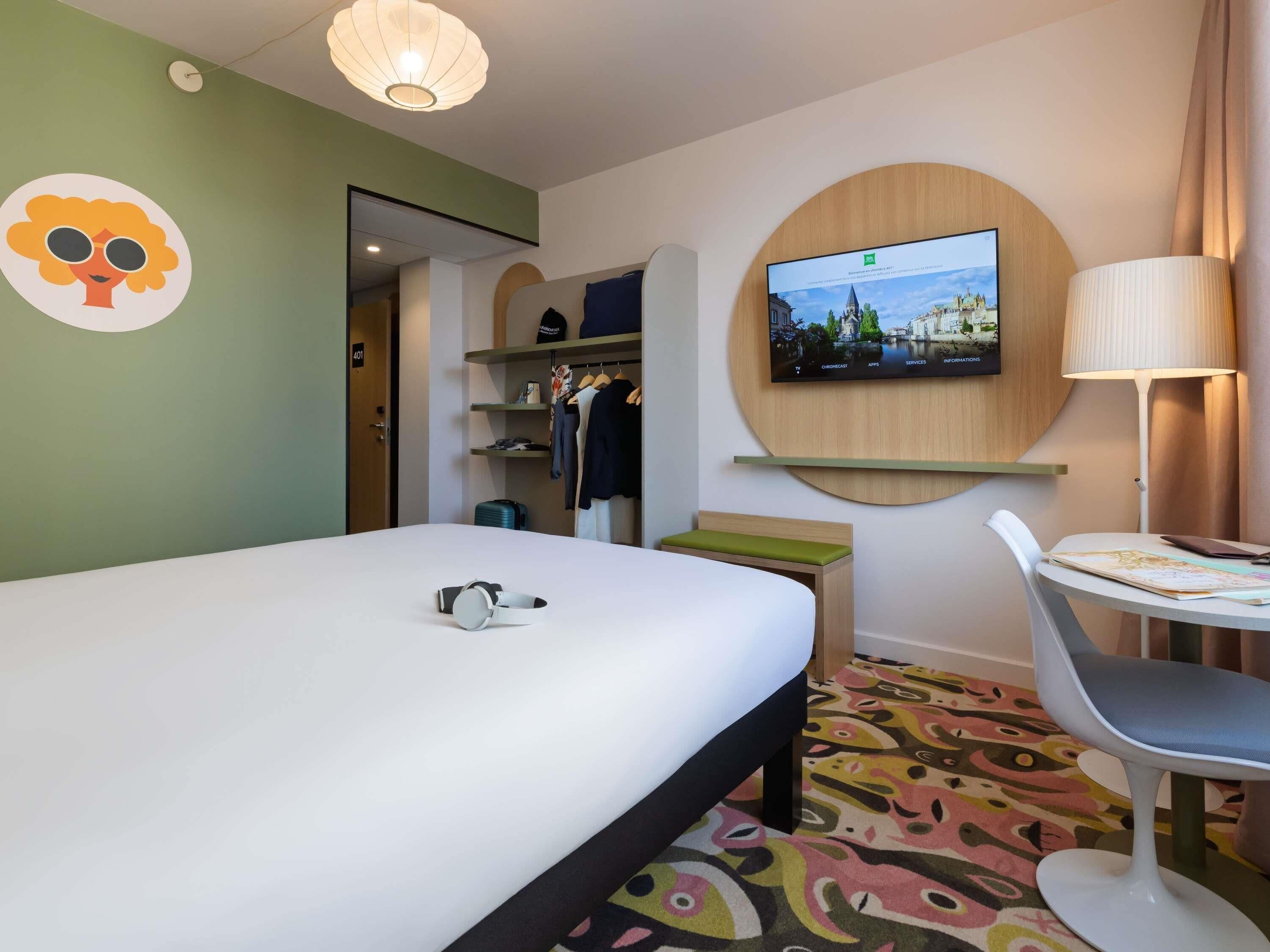 Quarto de hotel com uma cama, uma escrivaninha com cadeira, televisão e guarda-roupa.