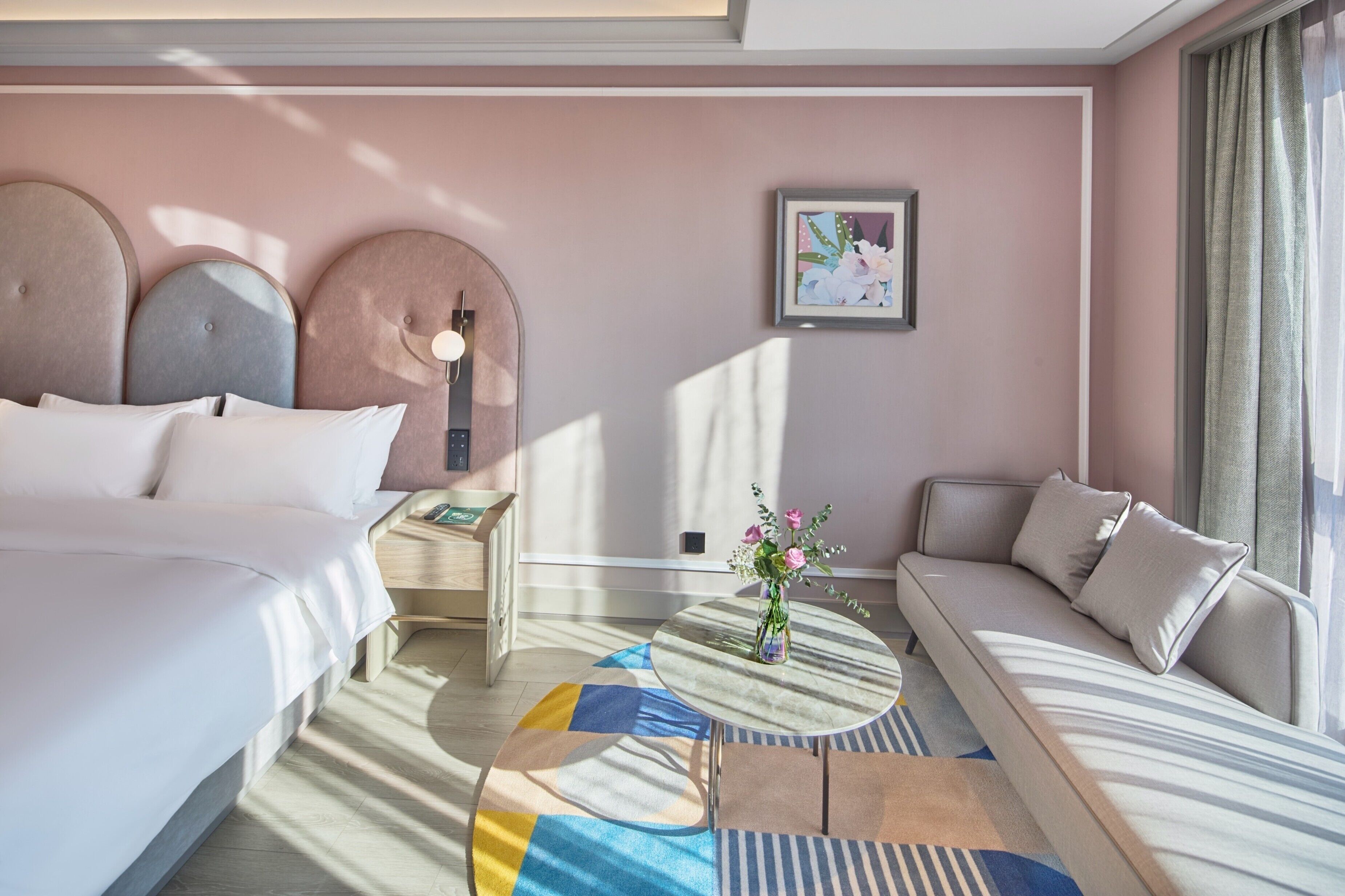 Quarto de hotel com cama, sofá, mesa redonda com um vaso de flores, pintura na parede e janela com cortinas.