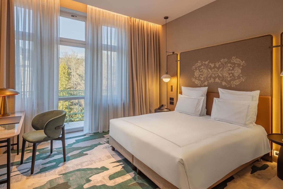Quarto de hotel com uma cama, uma cadeira, uma escrivaninha e vista para árvores pela janela.
