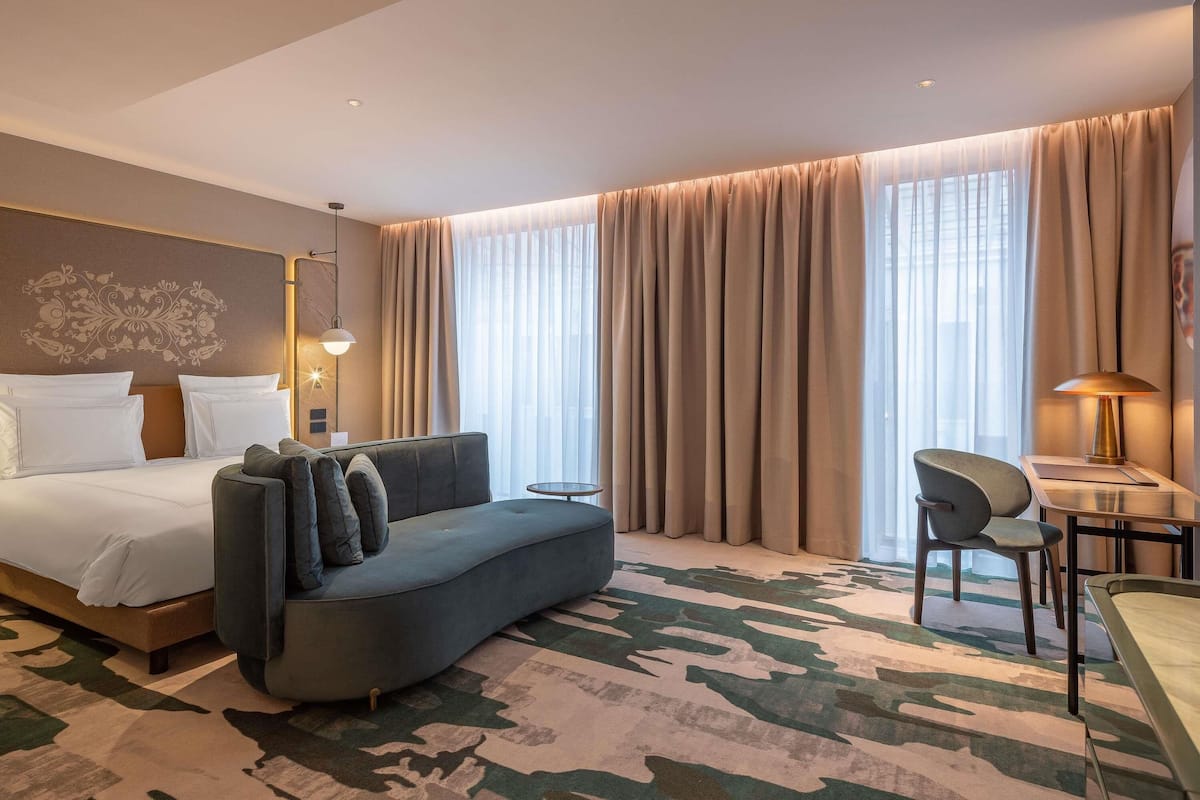 Quarto de hotel com cama, sofá, mesa pequena, cadeira e luminária.