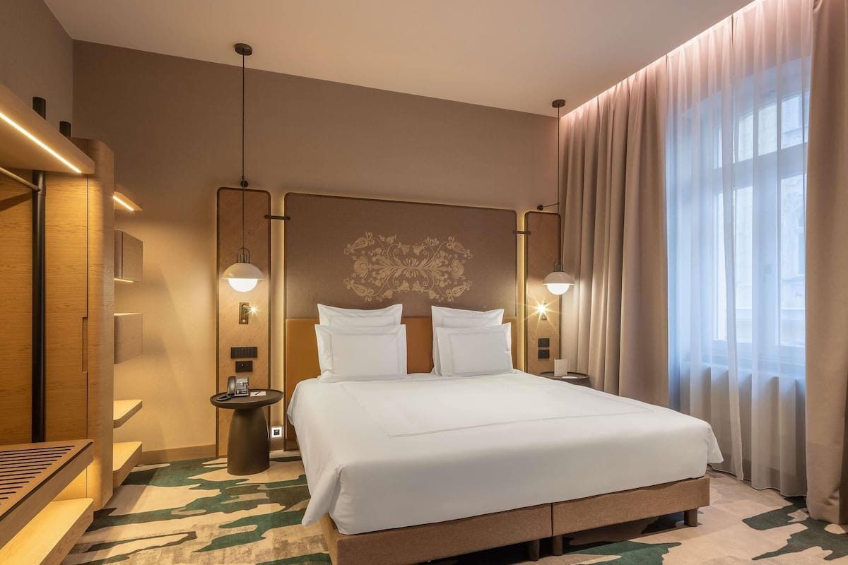 Quarto de hotel com cama, abajures, mesa de cabeceira, relógio na parede e janela com cortinas.