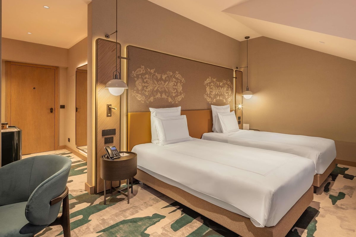 Quarto de hotel com duas camas, uma escrivaninha com telefone, uma cadeira e um tapete estampado.