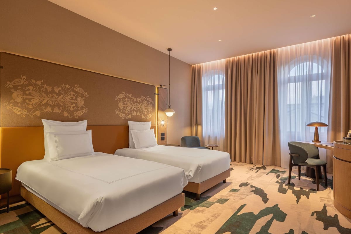 Quarto de hotel com duas camas, uma escrivaninha, uma cadeira, uma luminária e uma janela grande com cortinas.