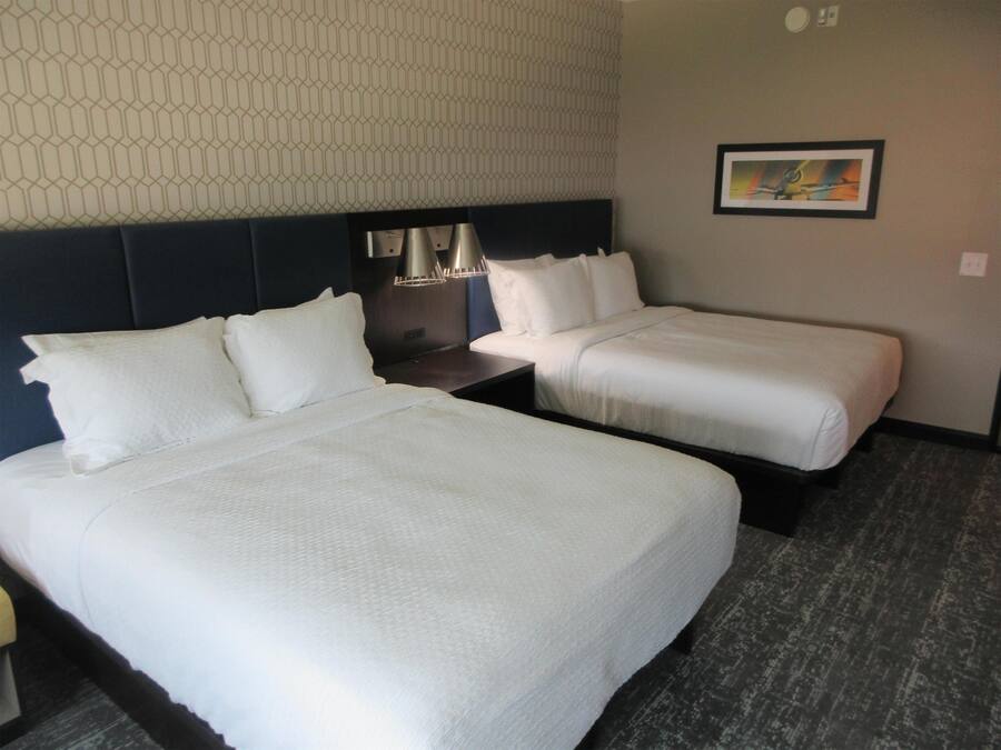 Quarto de hotel com duas camas, cabeceira, mesa de cabeceira com abajur e um quadro na parede.
