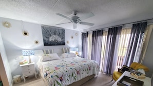 Quarto com cama, mesas de cabeceira, escrivaninha, ventilador de teto e cortinas.