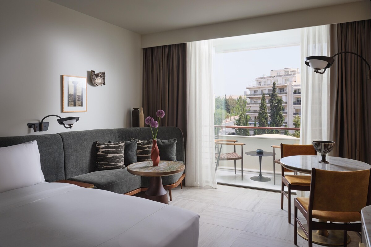Quarto de hotel com cama, sofá, mesa de jantar com cadeiras, um vaso com flores, luminária e vista para edifícios pela janela.