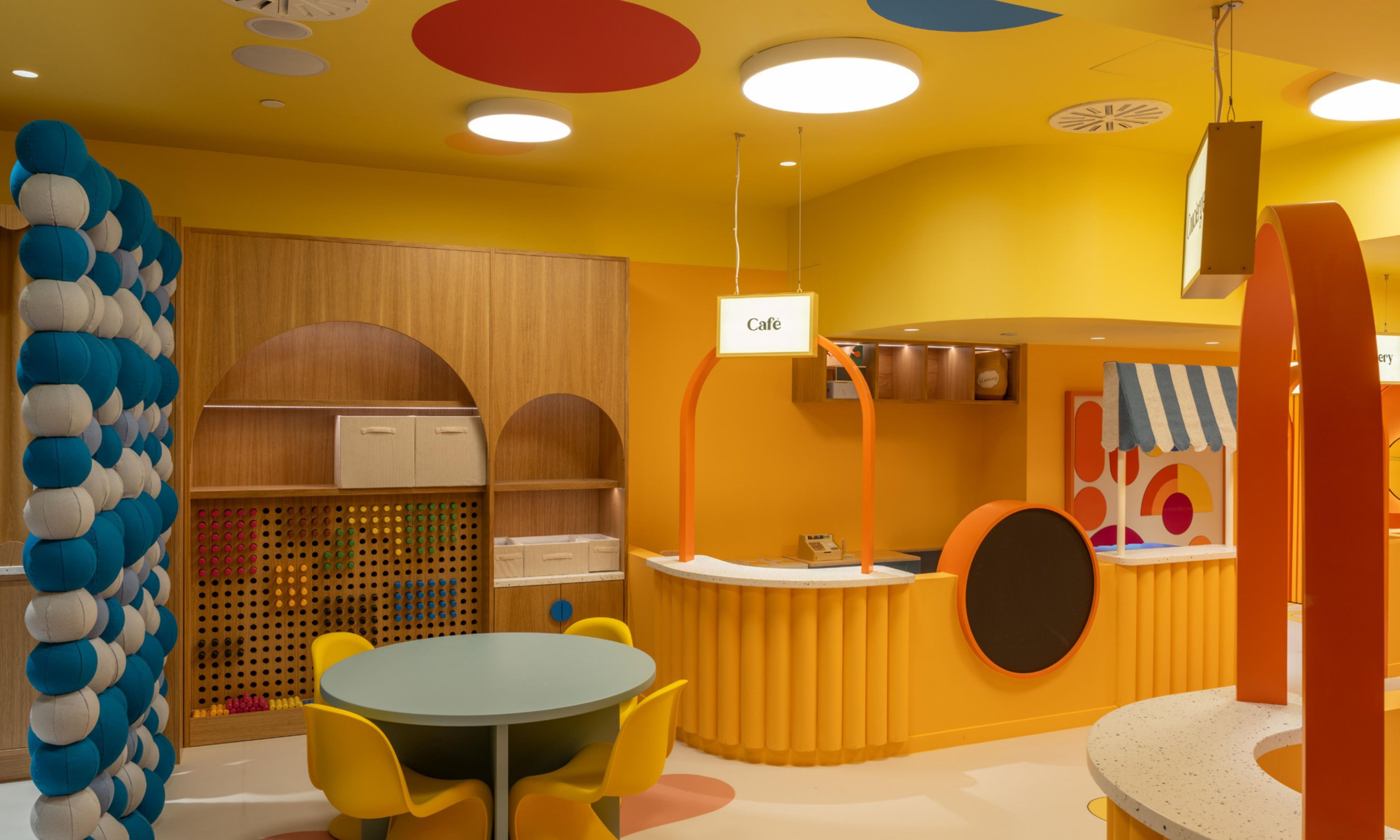 Uma área de recreação infantil, vibrante, com uma mesa redonda e cadeiras, um parquinho e um balcão identificado como 'Cafe'.
